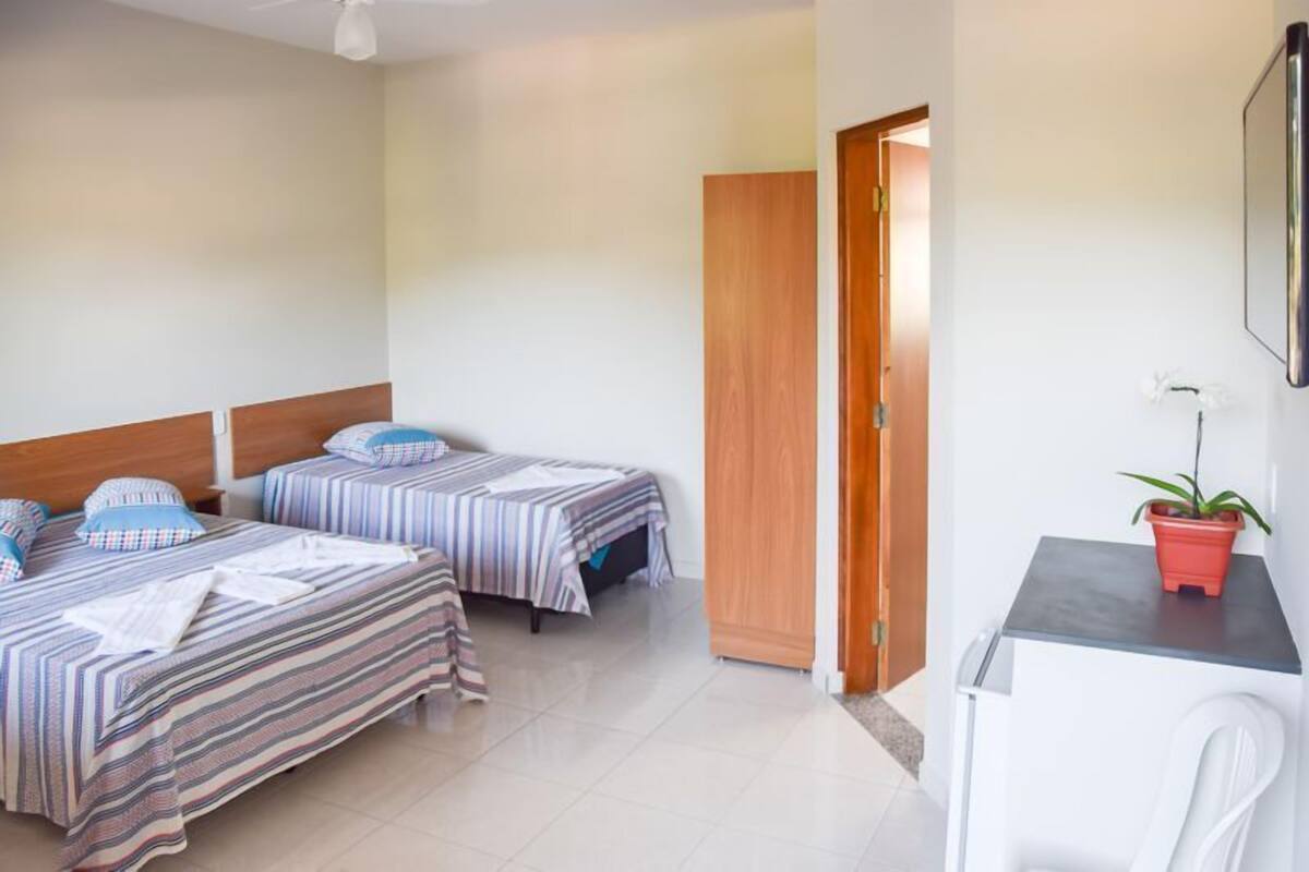 Quarto de hotel com duas camas, uma porta de madeira, uma escrivaninha com cadeira e um vaso de planta.
