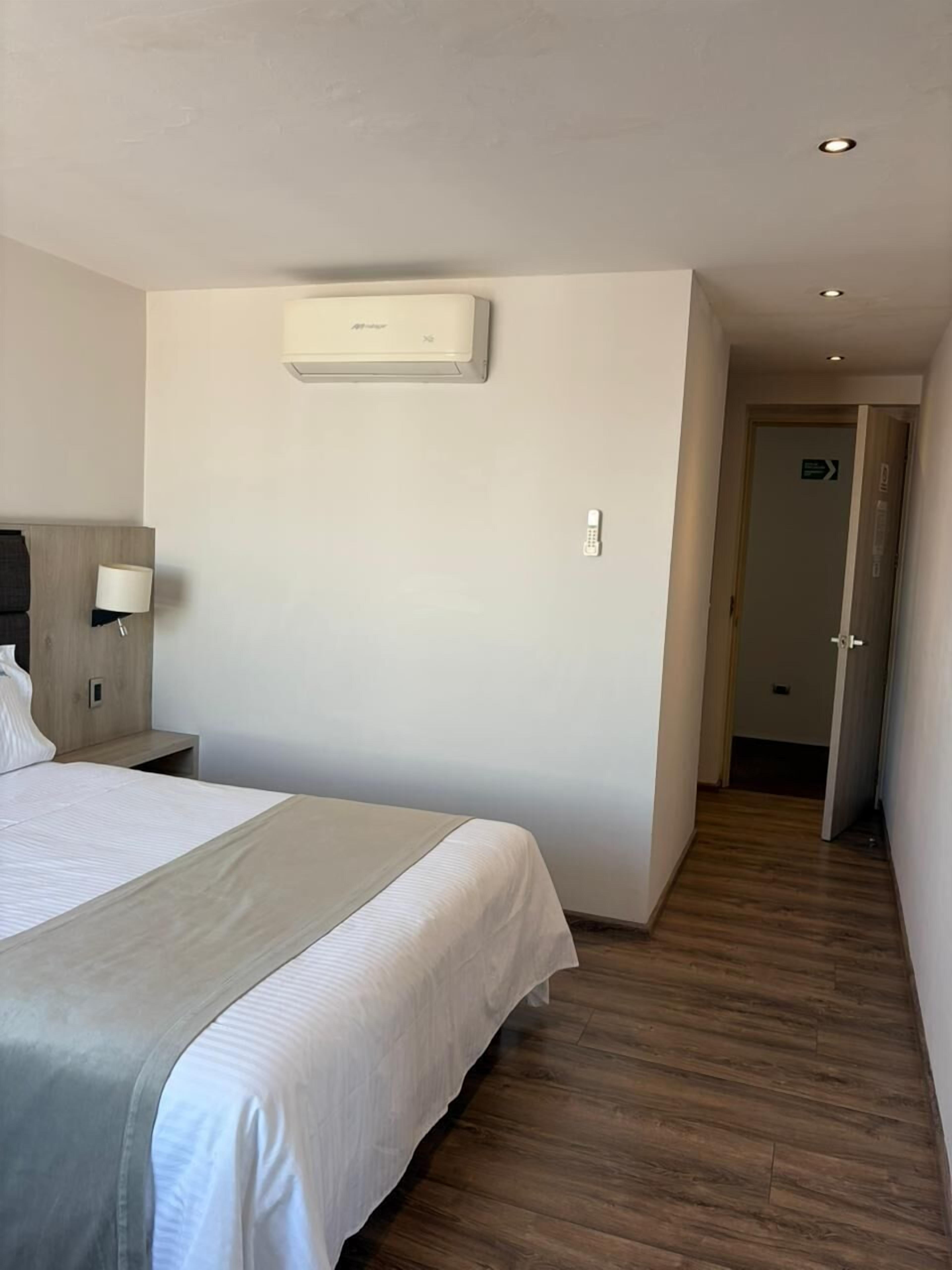Quarto de hotel com cama, mesa de cabeceira, ar-condicionado fixado na parede, porta e piso de madeira.