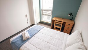 Uma cama bem arrumada com um edredom azul e branco, uma mesa de cabeceira de madeira com uma luminária e uma janela com vista para edifícios.