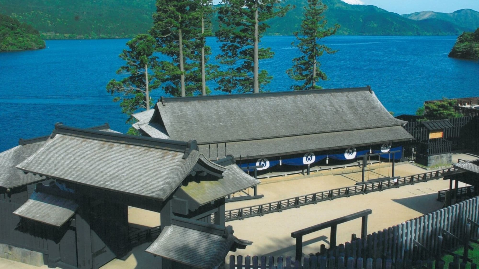 Um edifício japonês tradicional com telhado de palha, cercado por uma cerca de madeira, com vista para um lago e montanhas ao fundo.