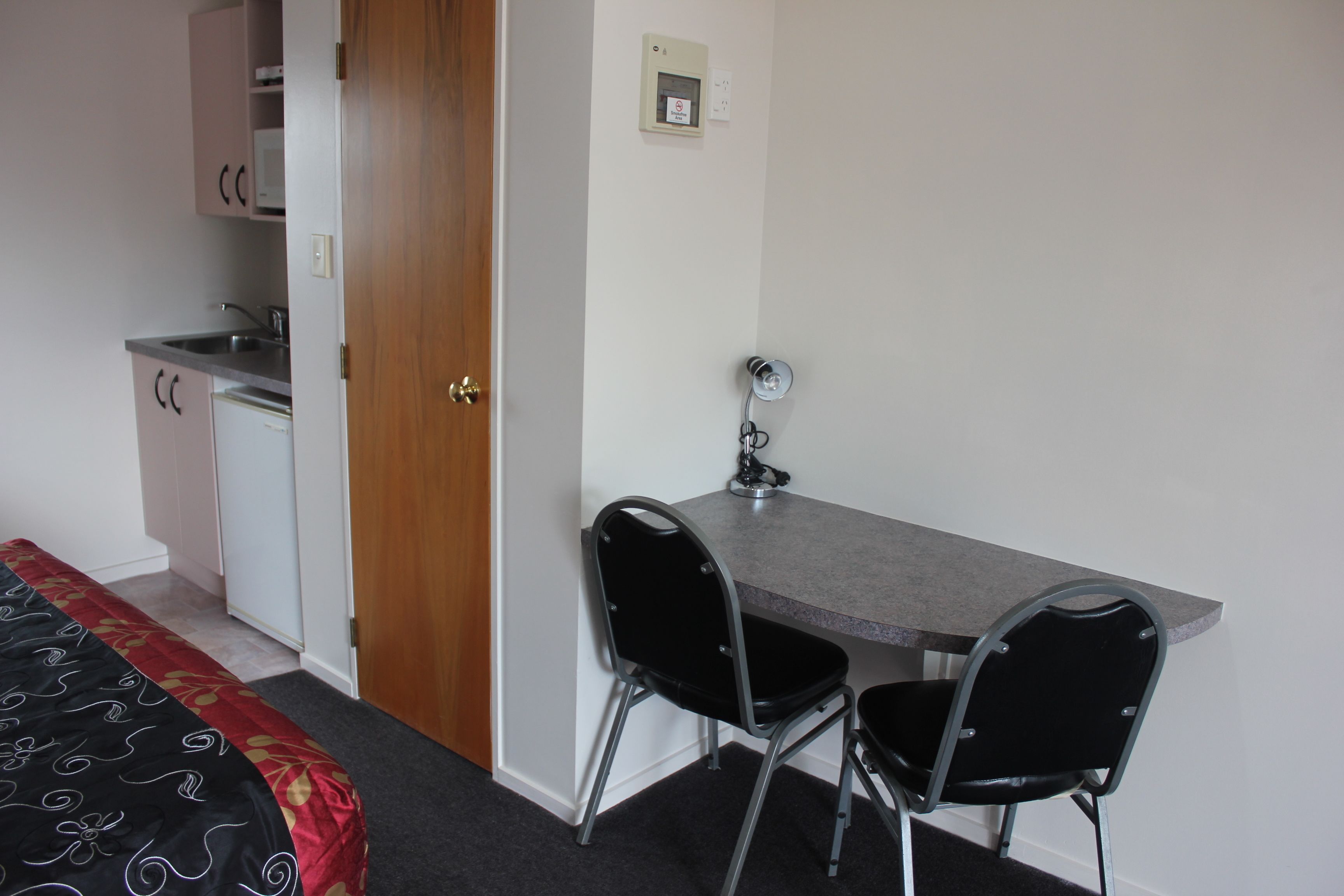 Quarto compacto com cozinha compacta, uma mesa com duas cadeiras e uma cama com colcha estampada.