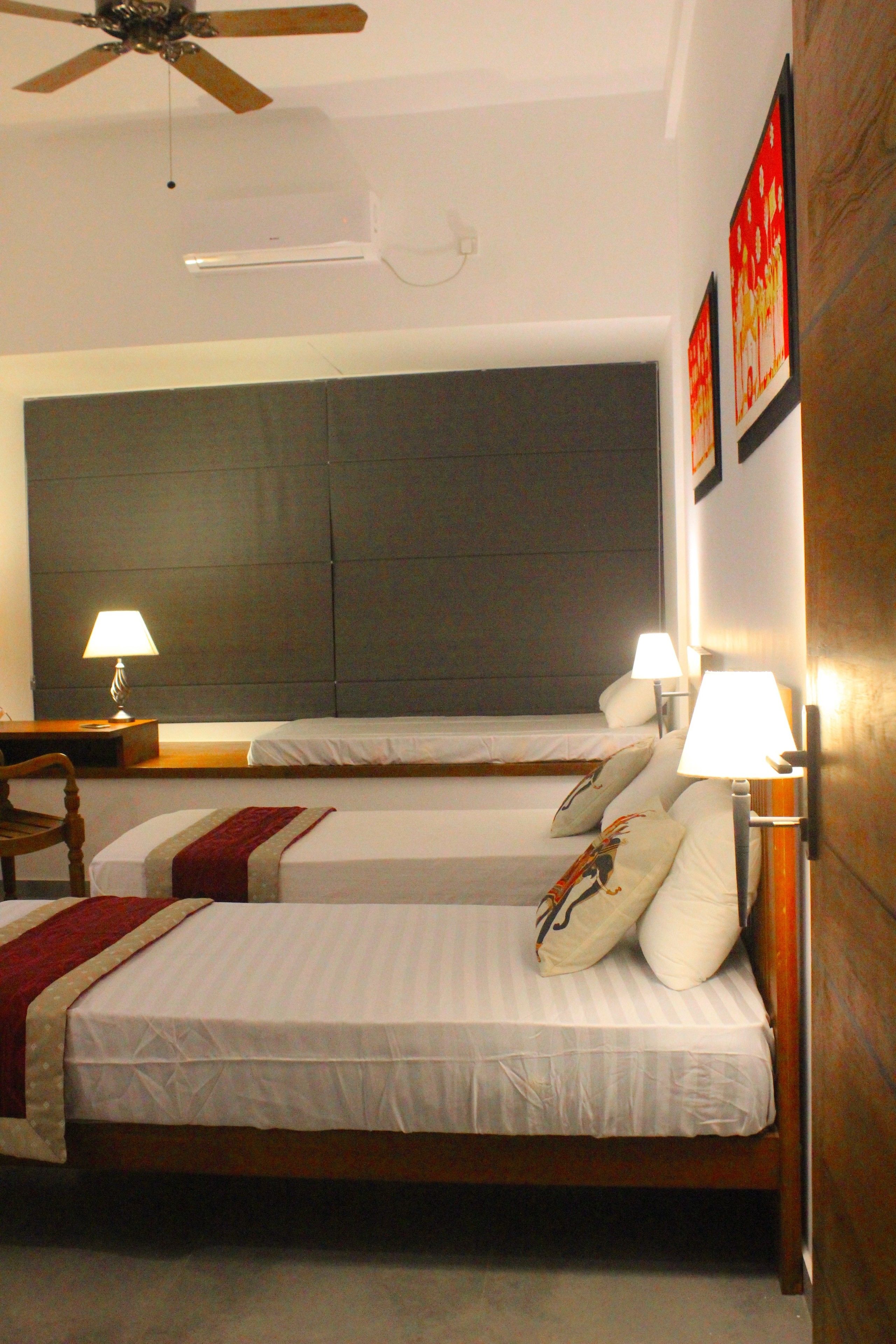 Quarto de hotel com duas camas de solteiro, cabeceira de madeira, ventoinha no teto e luminárias na parede.