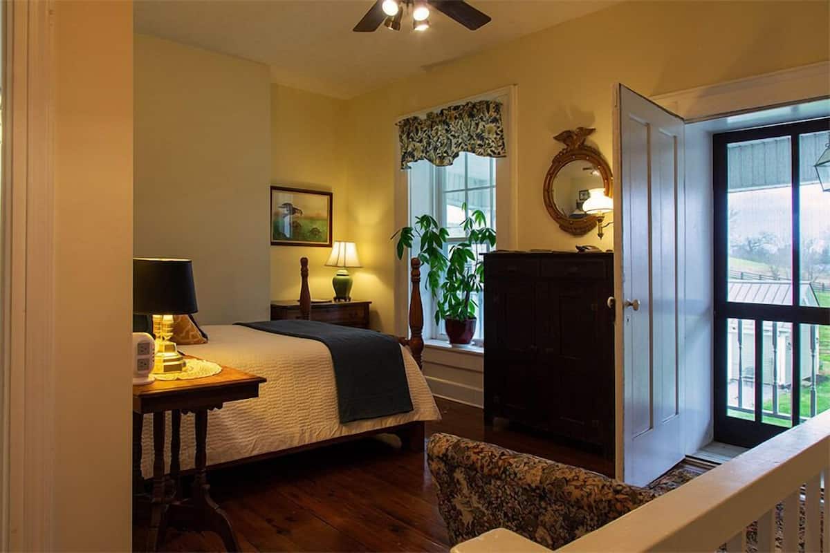 Quarto com cama, mesa de cabeceira com luminárias, uma cadeira, janela com cortinas, planta em vaso e porta que dá para uma varanda.