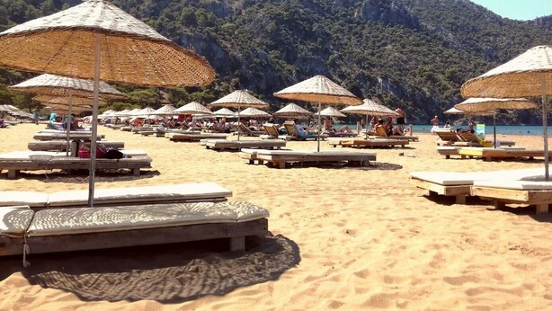 Uma praia com espreguiçadeiras e toldos de palha, com uma paisagem montanhosa ao fundo.