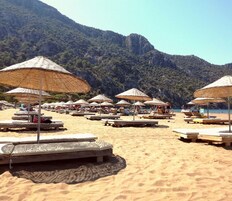 Uma praia com espreguiçadeiras e toldos de palha, com uma paisagem montanhosa ao fundo.