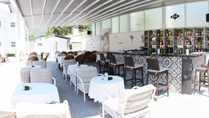 Uma área externa com mesas e cadeiras redondas, um bar com fundo estampado e vista para a paisagem ao redor.