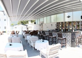 Uma área externa com mesas e cadeiras redondas, um bar com fundo estampado e vista para a paisagem ao redor.
