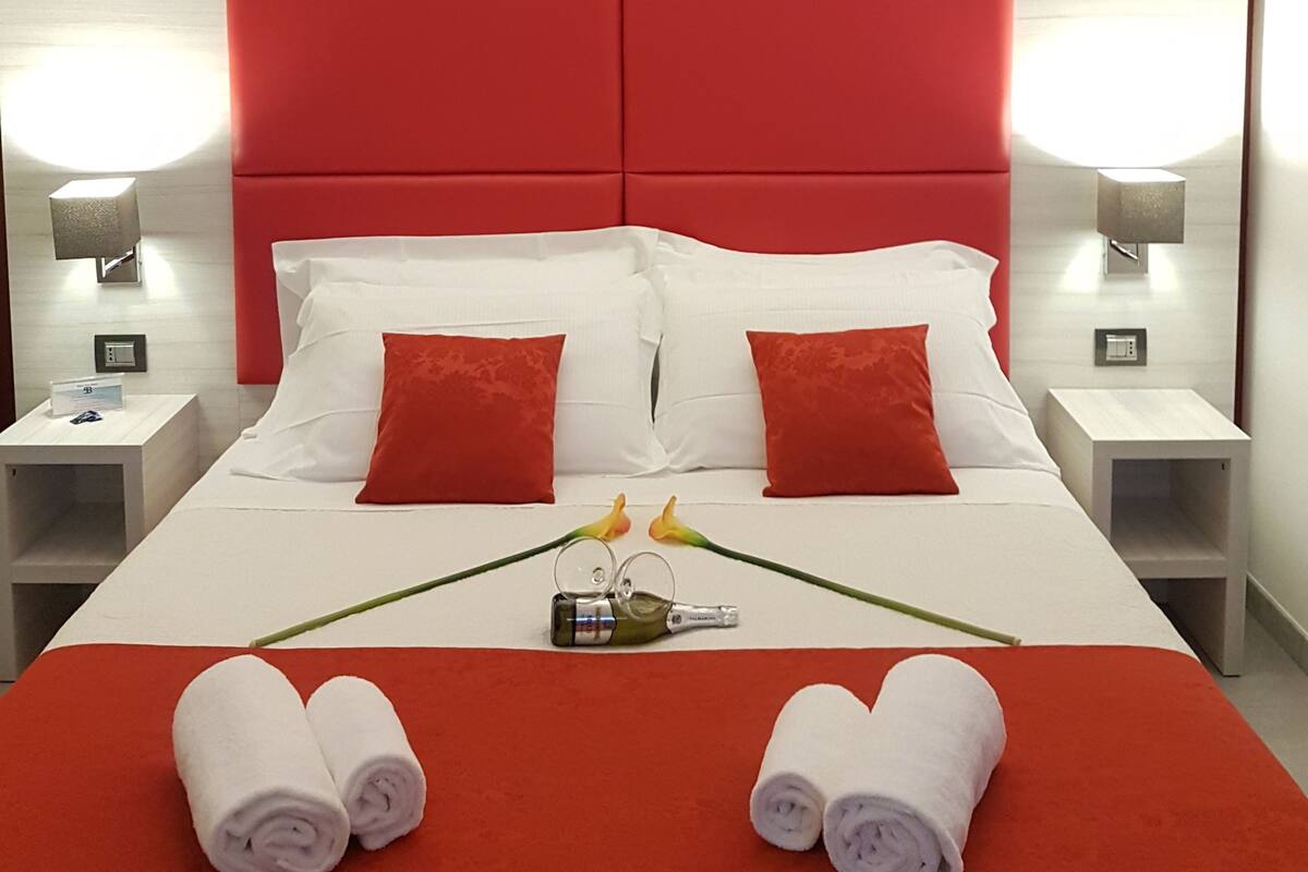 Quarto de hotel com uma cama grande, paredes em tons de vermelho e duas luminárias laterais.