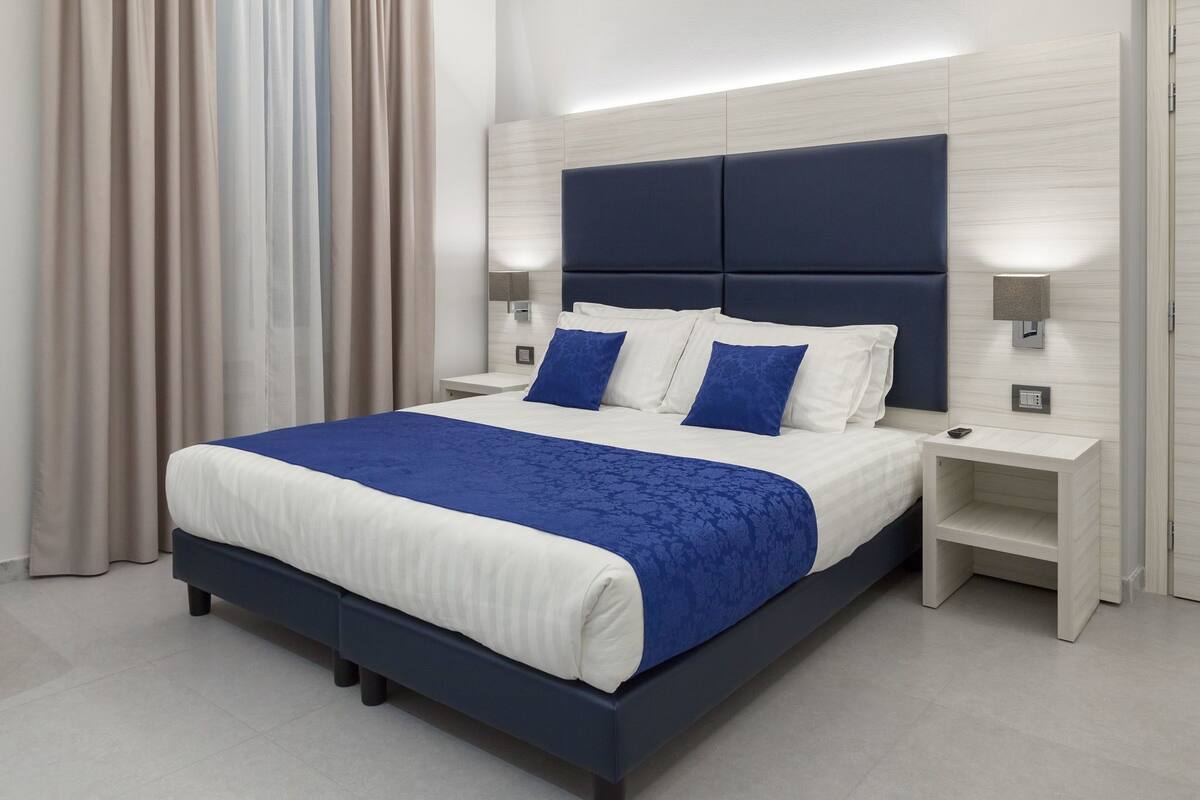 Quarto de hotel moderno com uma cama grande, duas mesas de cabeceira e uma mesinha de apoio.