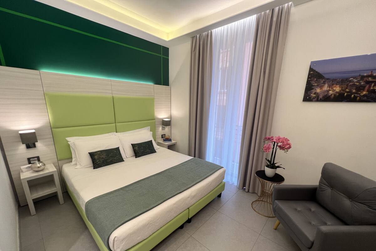 Quarto de hotel moderno com uma parede de destaque verde, uma cama grande com roupa de cama branca e verde, um sofá cinzento e vista para a cidade pela janela.