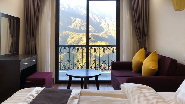 Quarto de hotel com uma cama, um sofá, uma mesinha e uma televisão. O quarto possui uma varanda com vista para as montanhas.