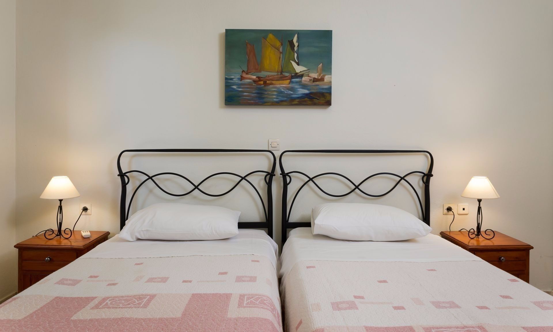 Uma cama de casal com colcha estampada, duas mesas de cabeceira com abajures e um quadro com barcos à vela na parede.