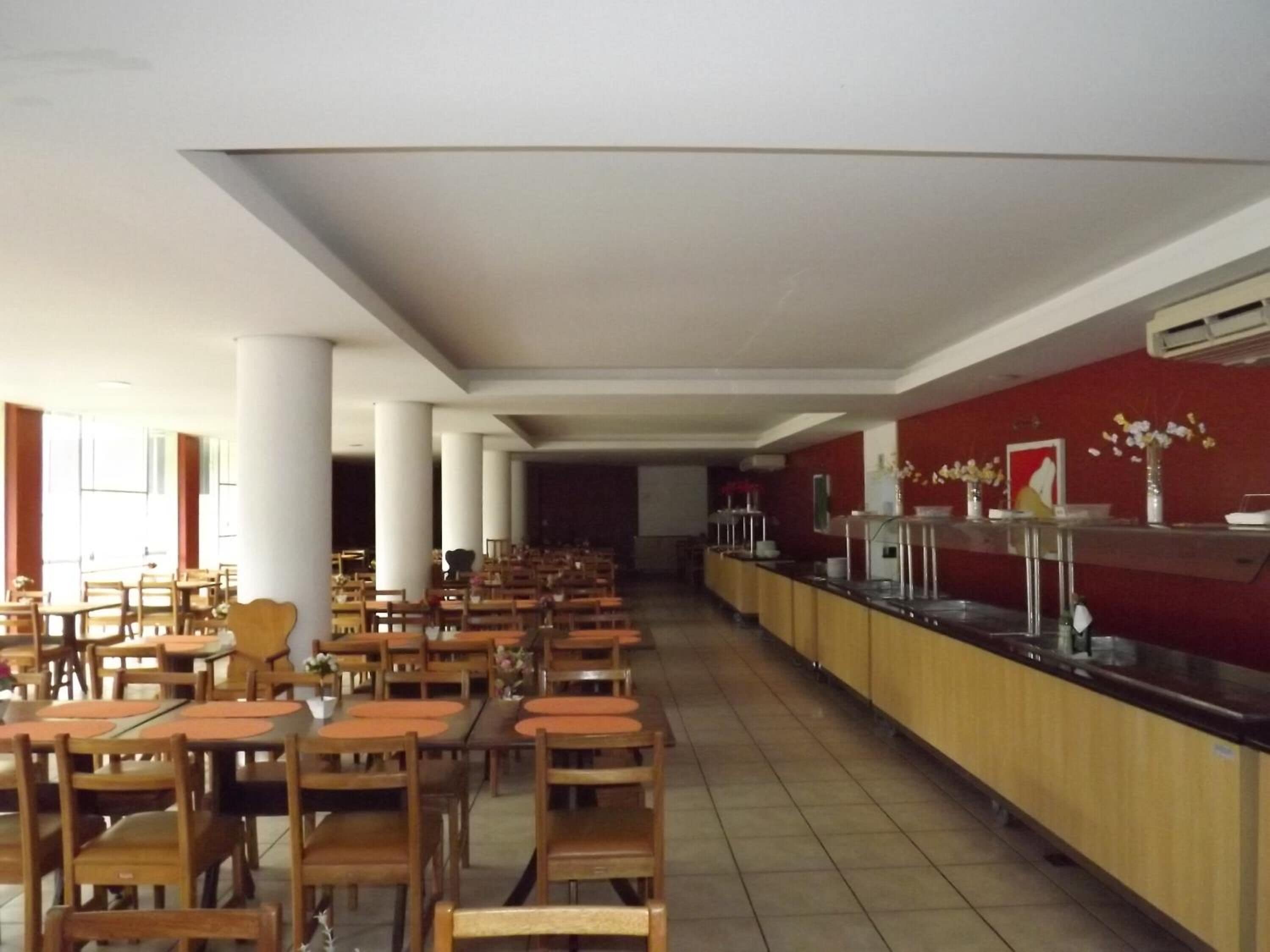 Um corredor longo com mesas e cadeiras de madeira, um balcão com parede vermelha e um aparelho de ar condicionado no teto.