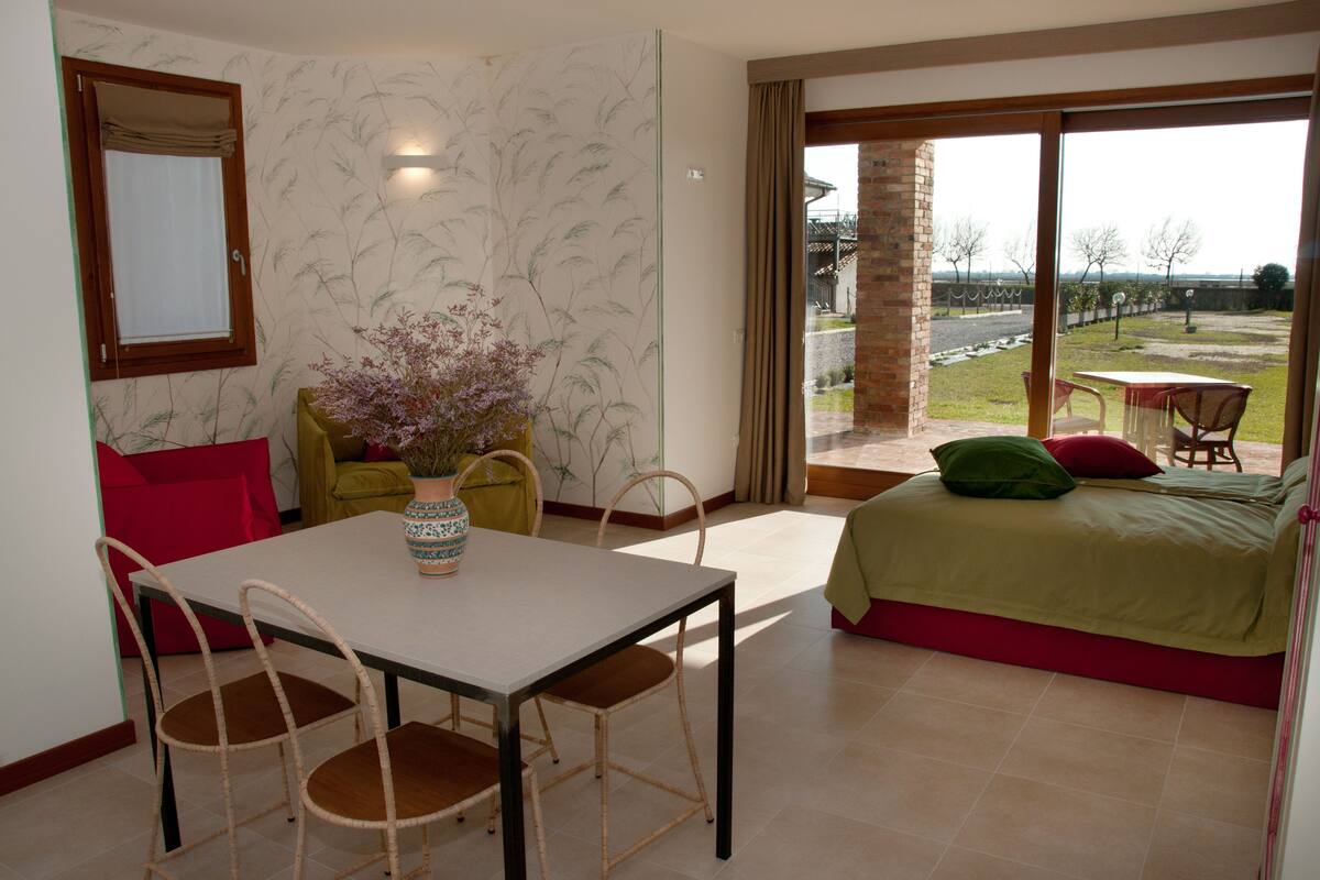 Quarto com mesa de jantar, cadeiras, sofá e cama. Há uma janela com vista para o exterior.