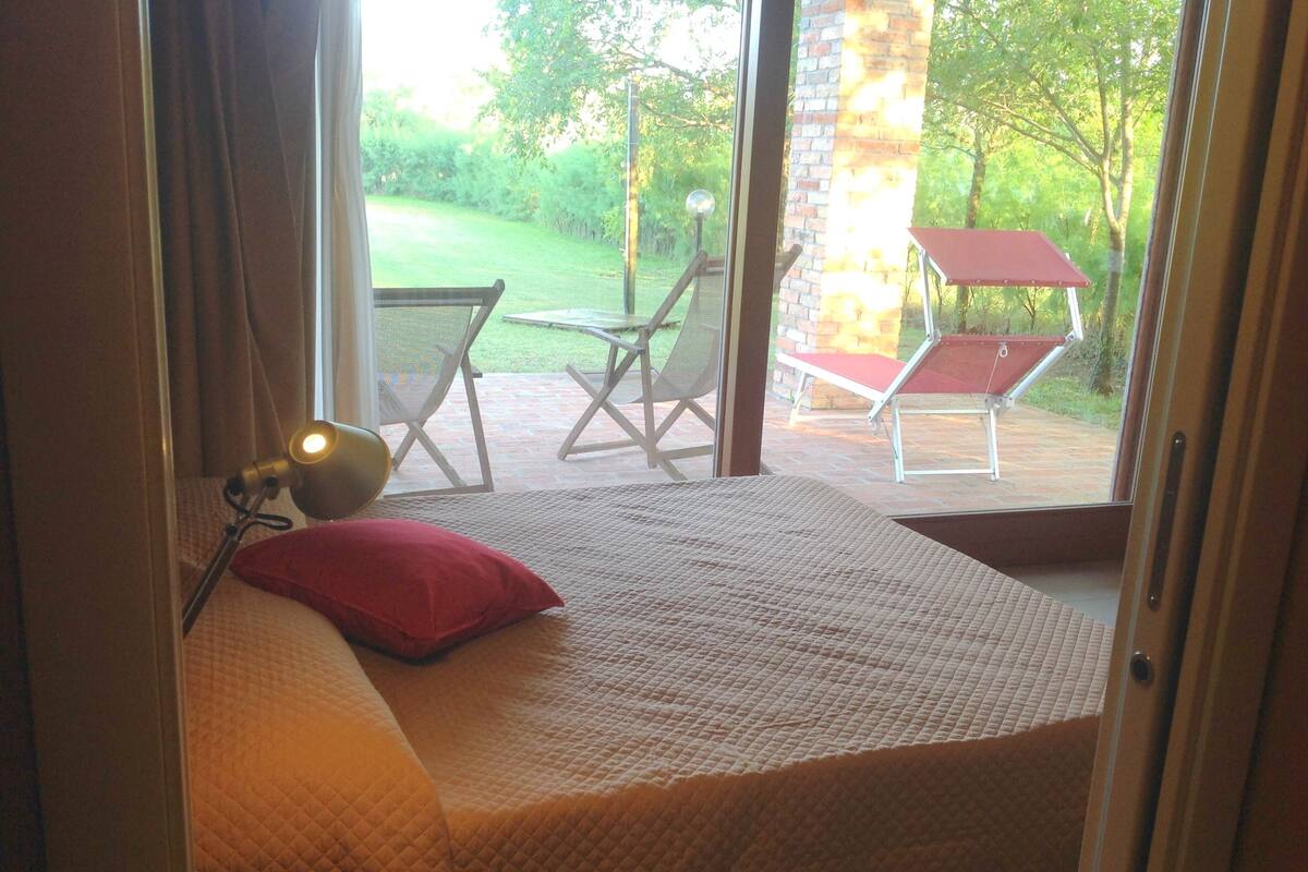 Quarto com cama, janela e vista para um pátio externo com mesa e cadeiras.