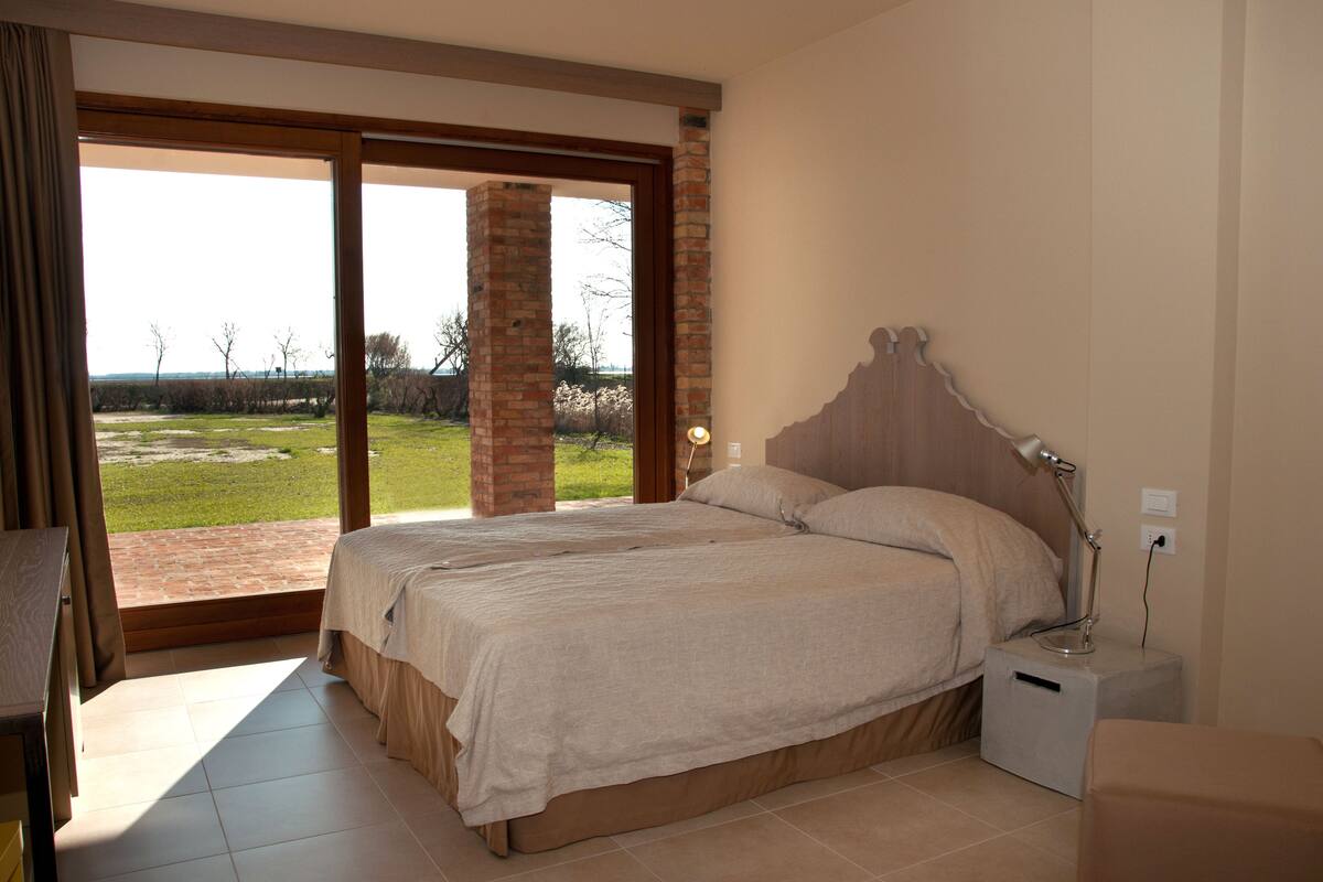 Quarto com uma cama grande, mesa de cabeceira, abajur e vista para uma área gramada através de uma porta de correr de vidro.