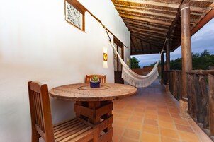 Uma mesa de madeira com uma cadeira, uma rede e um vaso de planta em um terraço com piso de azulejos e parede branca.