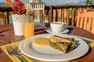 Uma mesa com um prato de comida, um copo de suco e uma xícara de café, montada ao ar livre com um cercado de madeira e vegetação ao fundo.
