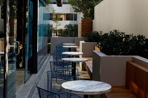 Uma área de estar moderna ao ar livre, com mesas redondas e cadeiras azuis, cercada por vegetação e fachada de um edifício.