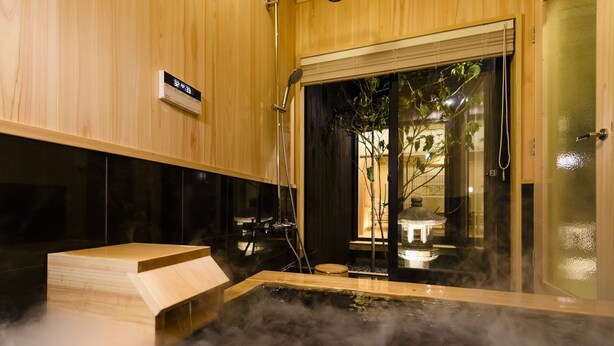 Uma onsen japonesa tradicional com interior de madeira, um banho de águas termais e vista para um templo através de uma porta de vidro.