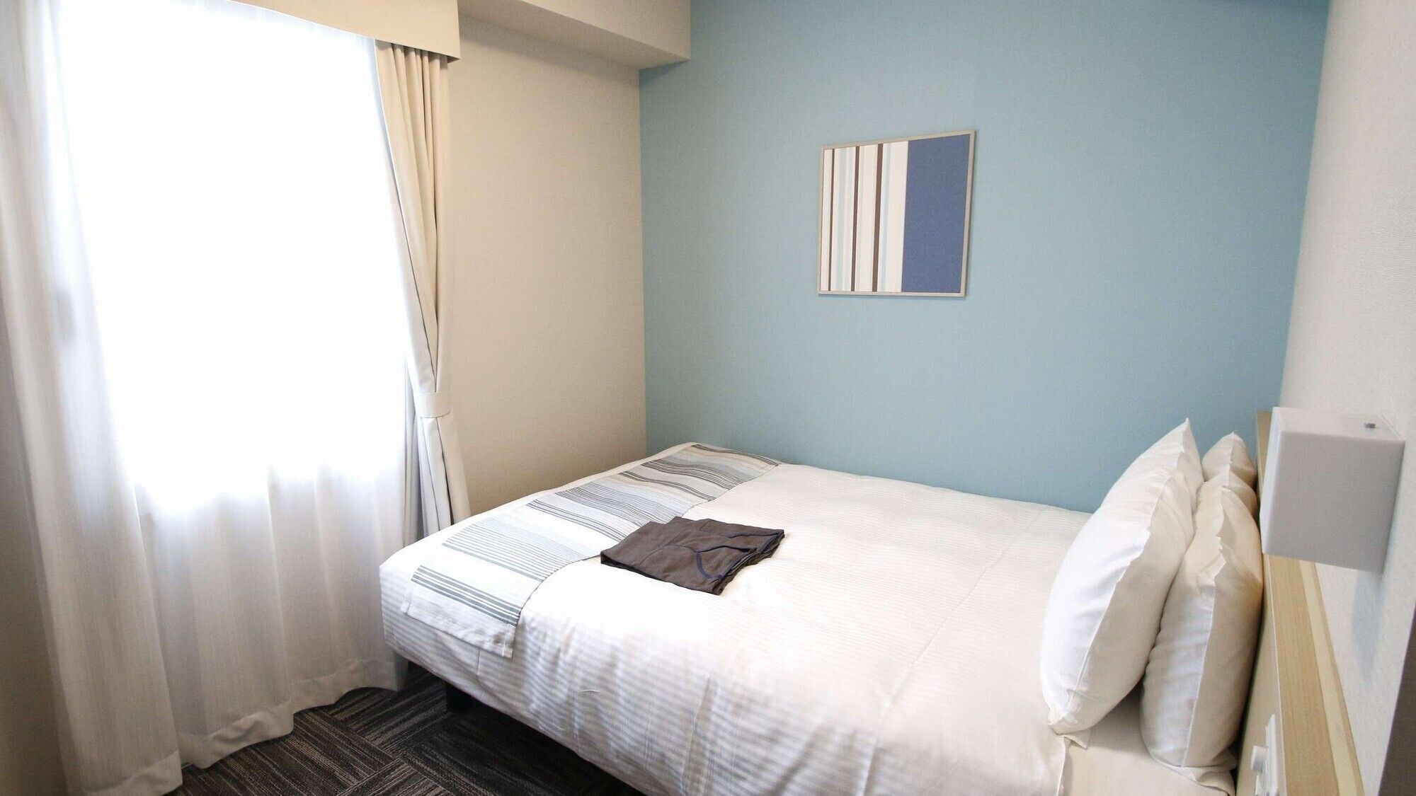 Quarto de hotel com cama, janela com cortinas, uma obra de arte na parede e uma mesa de cabeceira.