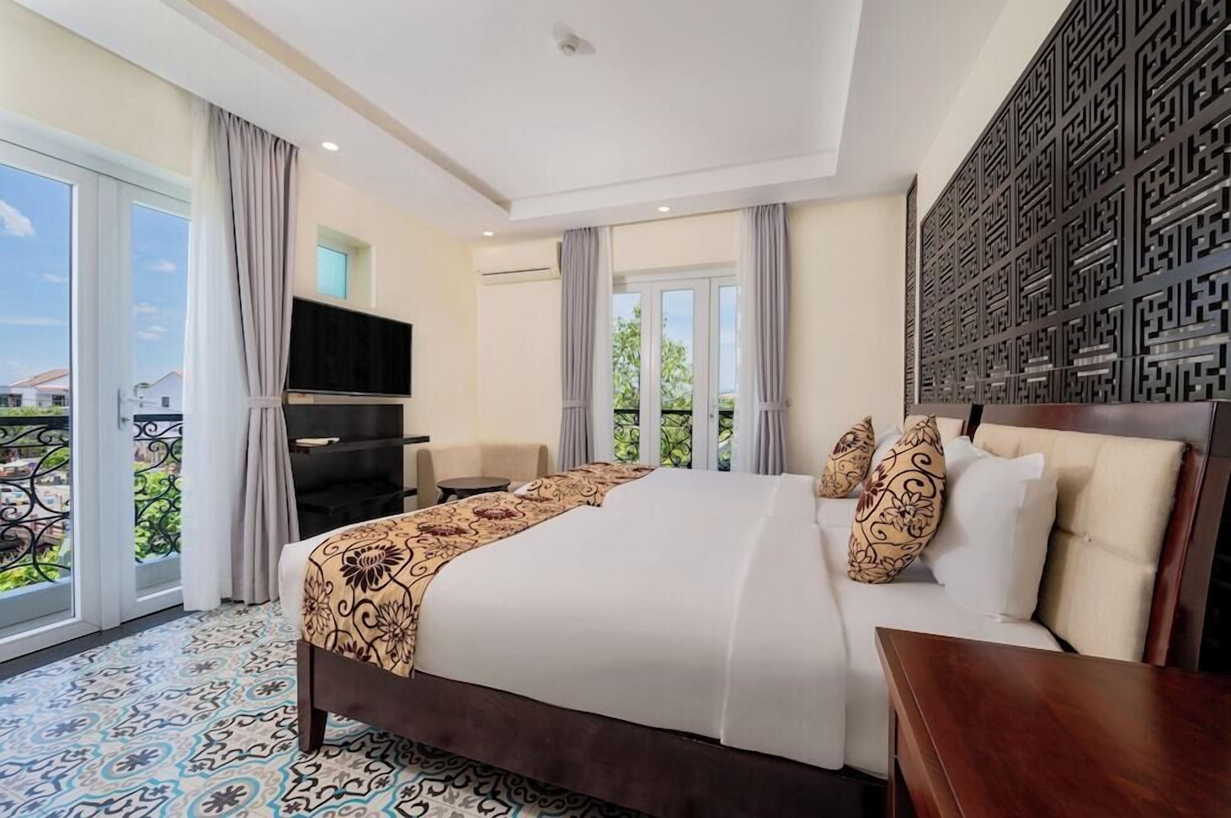 Quarto de hotel com uma cama grande, televisão, varanda com cortinas e piso estampado.