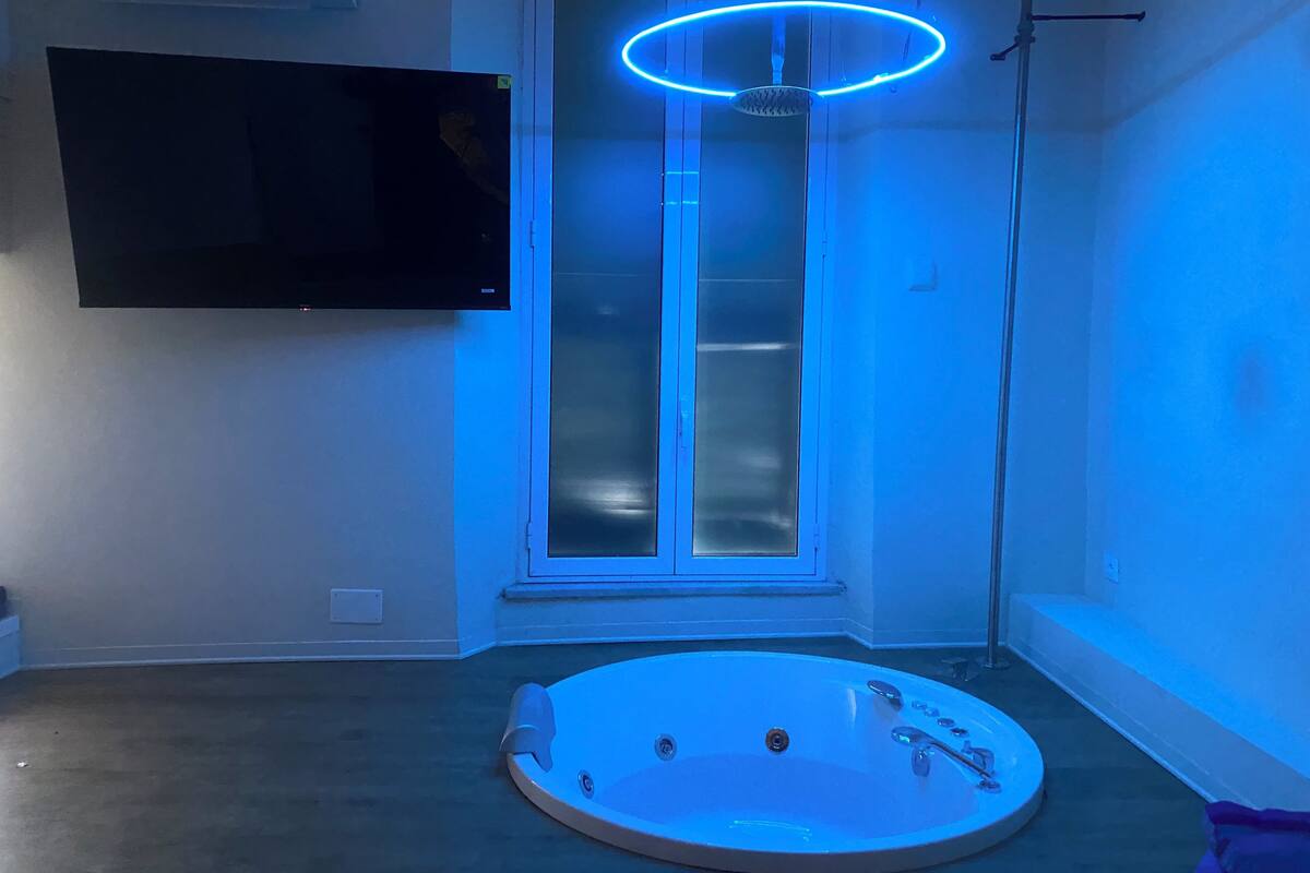 Quarto com uma janela grande, televisão montada e banheira circular com luminária azul em cima.