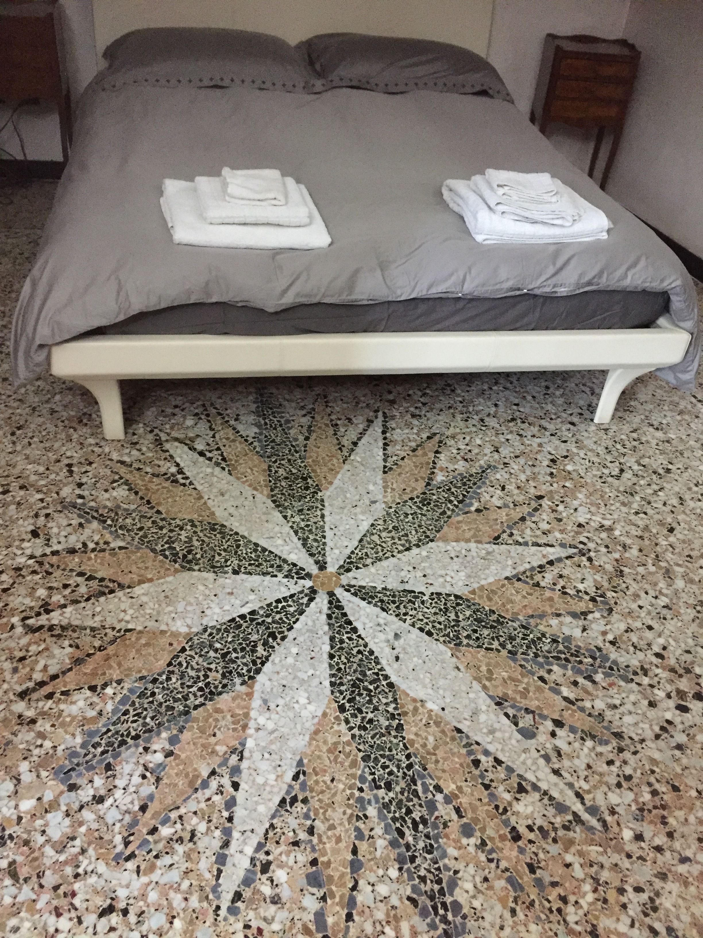 Cama com colcha cinza e duas toalhas brancas dobradas no encosto. O chão apresenta um desenho de mosaico em formato de estrela.