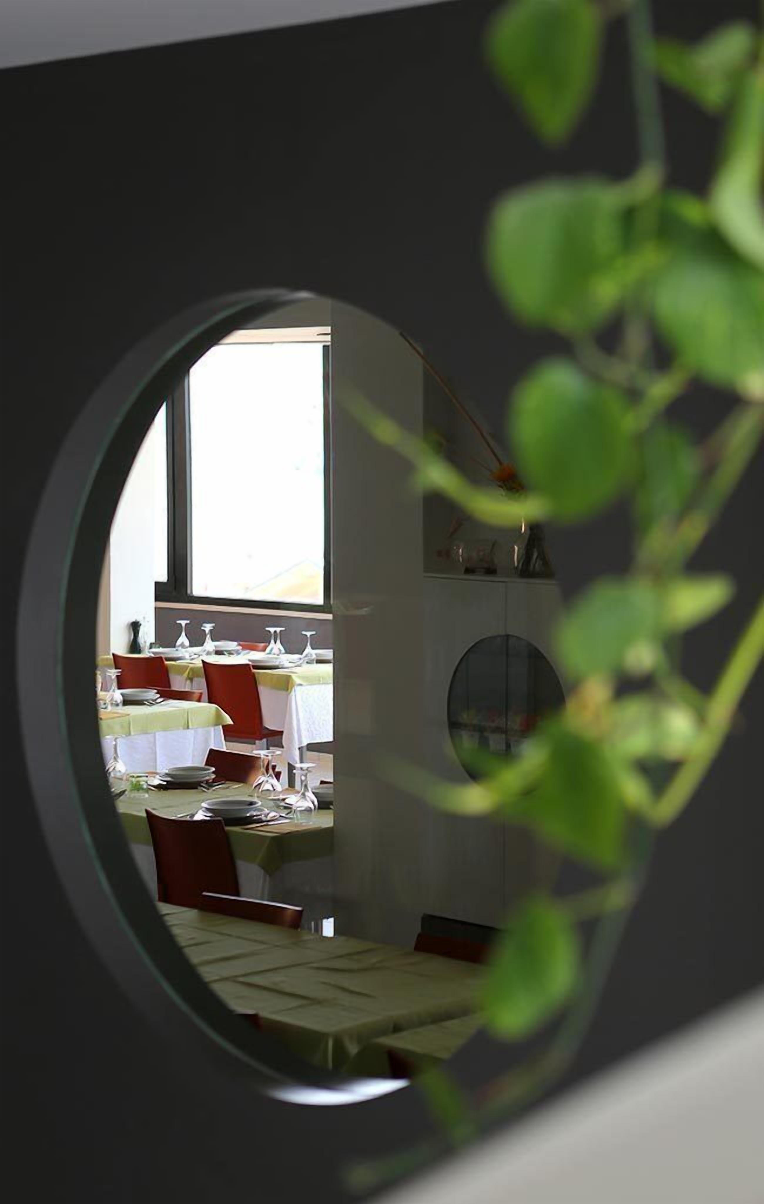 Quarto com uma janela grande, um espelho redondo refletindo o ambiente, e uma planta em primeiro plano.