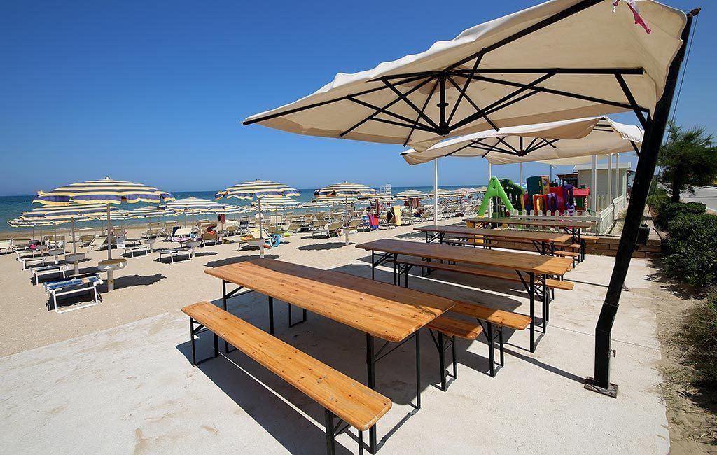 Um café à beira-mar com mesas e bancos de madeira ao ar livre, e vários guarda-sóis de praia.