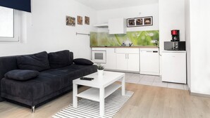 Um espaço de estar moderno com sofá preto, mesa de centro branca e área de cozinha com armários e eletrodomésticos brancos.