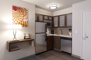 Cozinha compacta com geladeira, micro-ondas e lava-louças. Há um armário de madeira com porta de vidro, uma bancada com cafeteira e uma prateleira na parede com uma luminária e um quadro emoldurado.