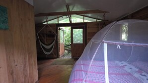 Uma cabana rústica de madeira com rede, cama com mosquiteiro e um alpendre de madeira.