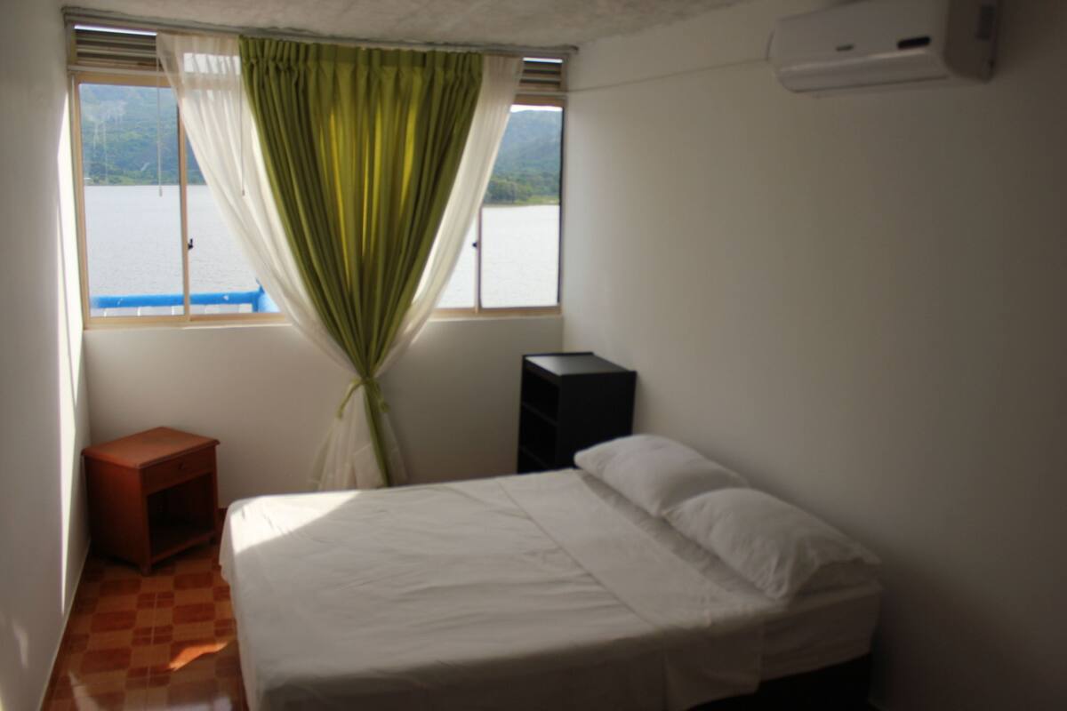 Quarto pequeno e simples com cama, mesa de cabeceira, janela com cortinas e ar condicionado fixado na parede.