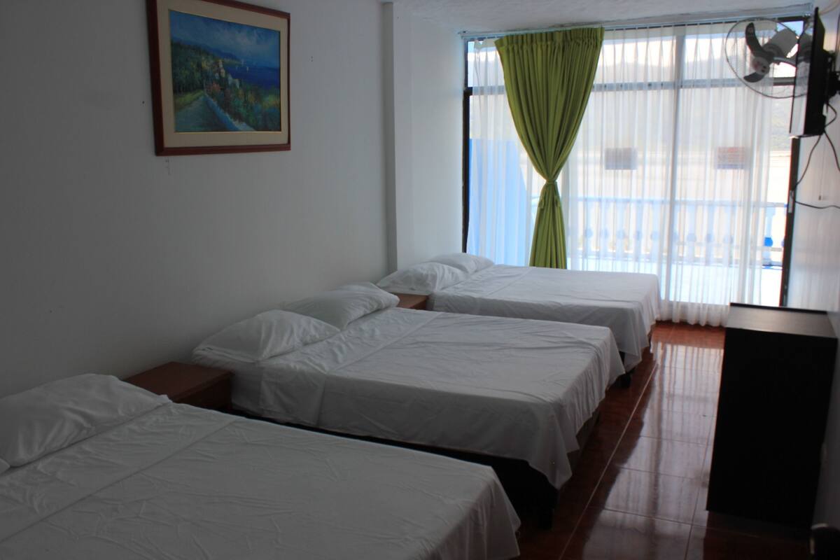 Quarto de hotel com três camas de solteiro, um quadro na parede, uma janela com cortinas e uma mesa com abajur.