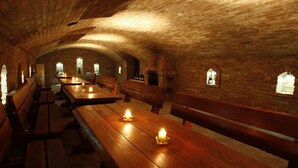 Um interior com pouca luz, paredes de tijolo, bancos e mesas de madeira, pequenas janelas e uma lareira.