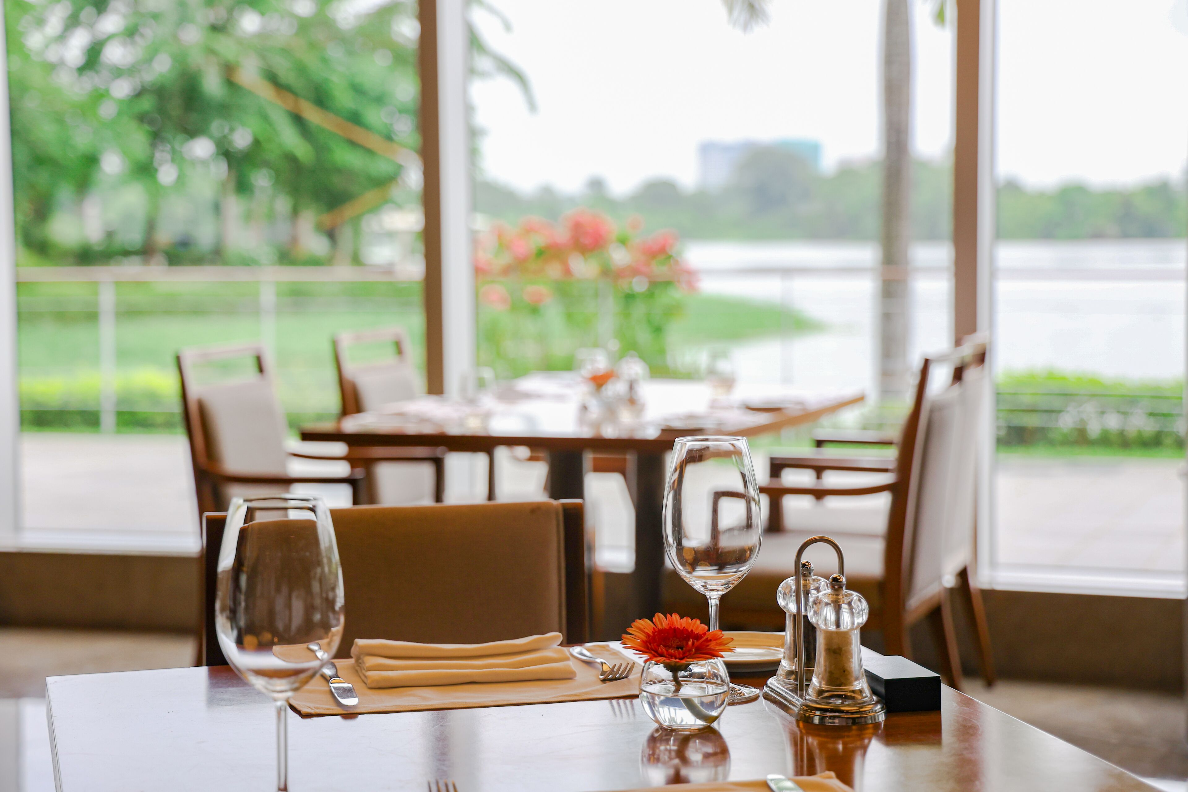 Uma mesa de jantar posta com uma taça de vinho, um pequeno vaso com uma flor e um saleiro e pimenteiro. A mesa está posicionada perto de grandes janelas com vista para um corpo d'água e vegetação.