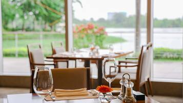 Uma mesa de jantar posta com uma taça de vinho, um pequeno vaso com uma flor e um saleiro e pimenteiro. A mesa está posicionada perto de grandes janelas com vista para um corpo d'água e vegetação.