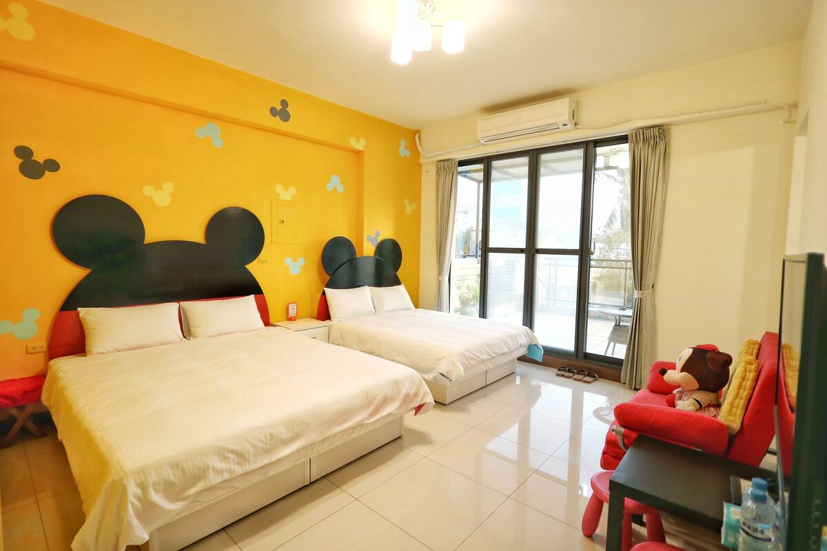 Quarto de hotel com duas camas, parede com tema do Mickey Mouse, uma cadeira vermelha e uma janela grande.
