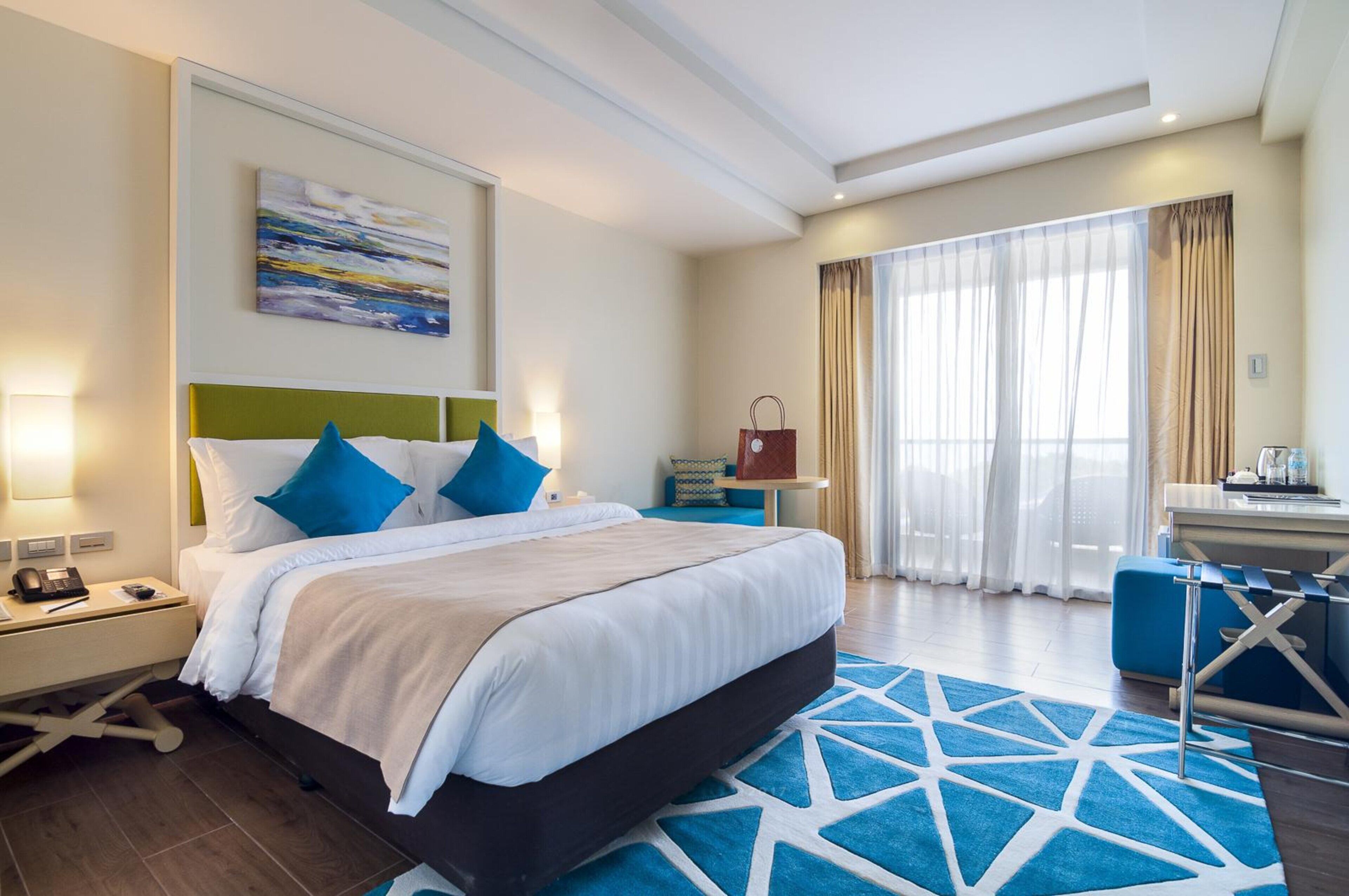 Quarto de hotel com uma cama grande, escrivaninha, cadeira, mesa de cabeceira, luminária, um quadro na parede e uma janela com cortinas.