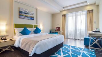 Quarto de hotel com uma cama grande, escrivaninha, cadeira, mesa de cabeceira, luminária, um quadro na parede e uma janela com cortinas.