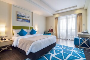 Quarto de hotel com uma cama grande, escrivaninha, cadeira, mesa de cabeceira, luminária, um quadro na parede e uma janela com cortinas.