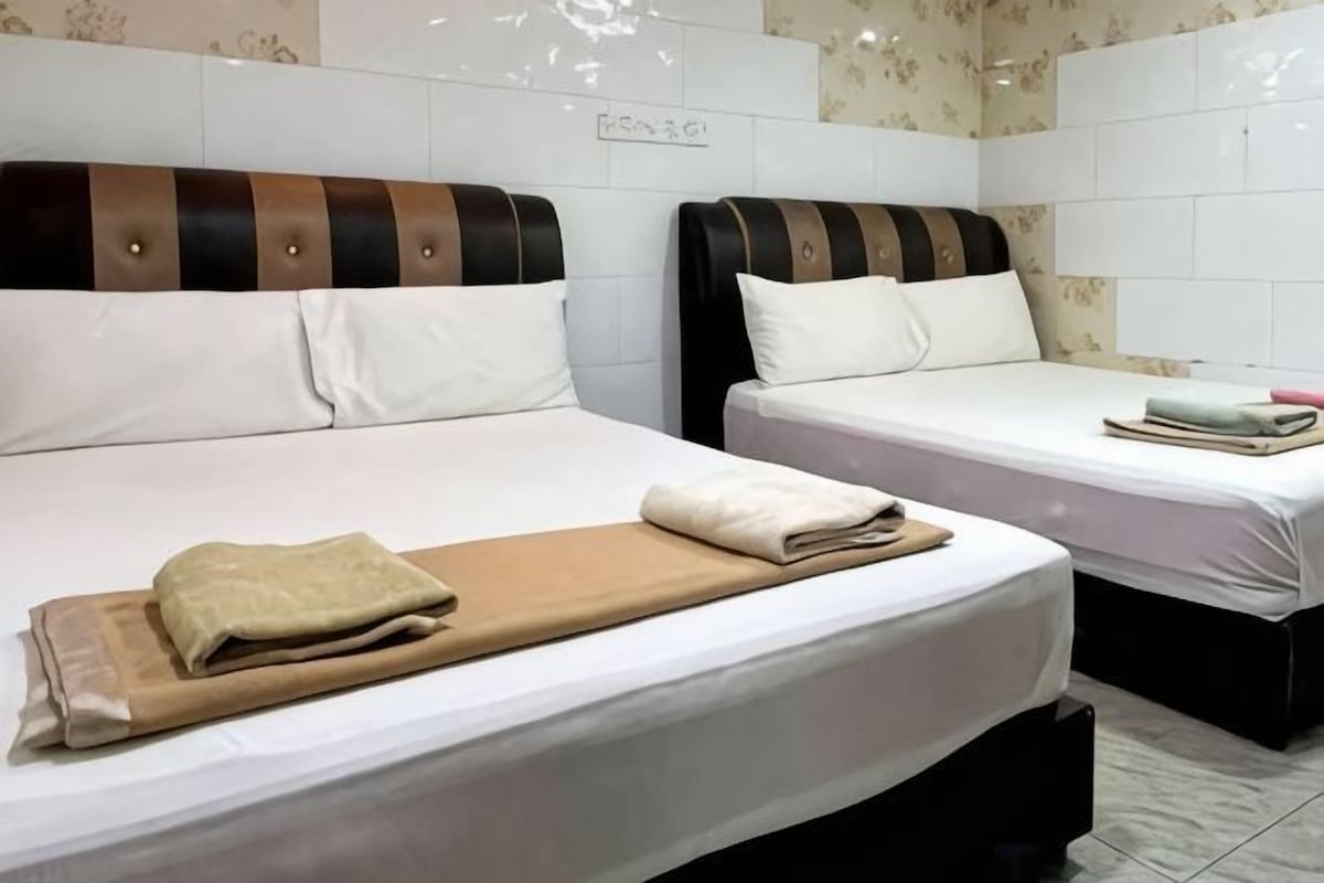 Duas camas de solteiro com roupa de cama branca, um cobertor marrom e uma pequena mesa de cabeceira com um livro e um objeto rosa.