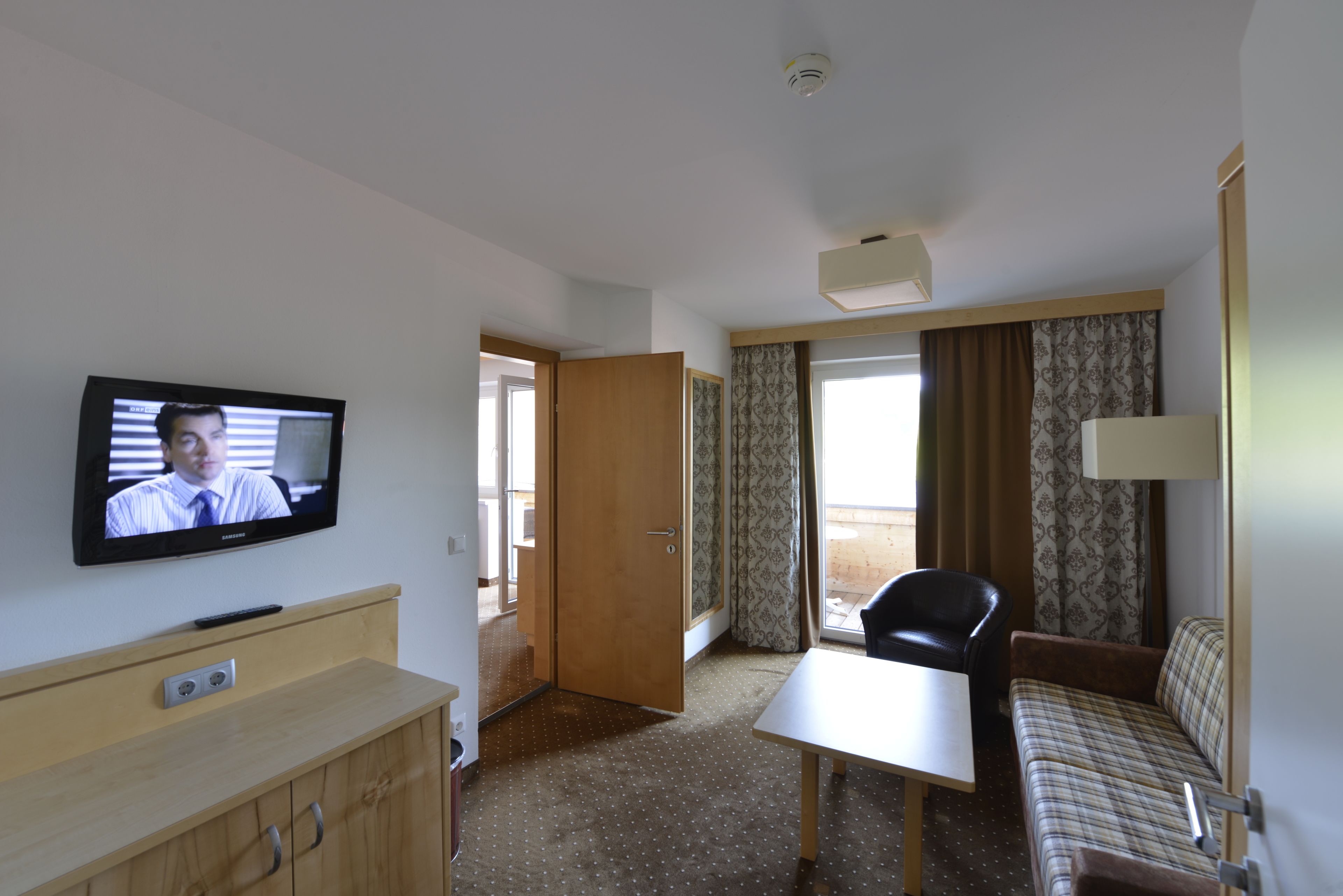 Quarto de hotel com televisão, sofá, mesa e porta que dá para uma varanda.