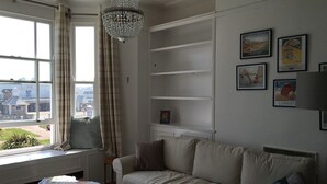 Uma sala de estar com sofá, um assento junto à janela e um candelabro. Há quadros nas paredes e um radiador abaixo do sofá.