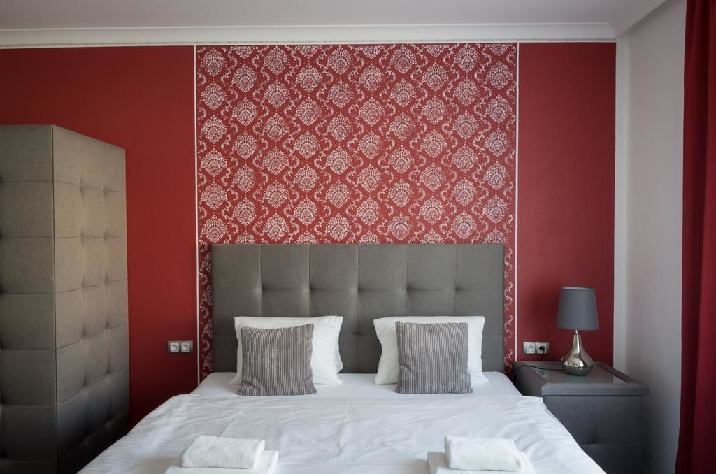 Quarto de hotel com cabeceira estofada, uma cama grande com roupa de cama branca, duas almofadas, uma mesa de cabeceira com abajur e papel de parede vermelho estampado.