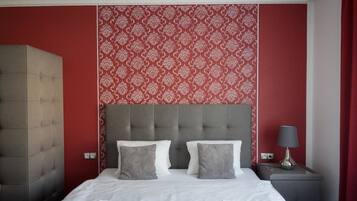 Quarto de hotel com cabeceira estofada, uma cama grande com roupa de cama branca, duas almofadas, uma mesa de cabeceira com abajur e papel de parede vermelho estampado.