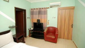 Quarto com uma cama, televisão sobre um suporte, poltrona vermelha, porta de madeira e aparelho de ar condicionado na parede.