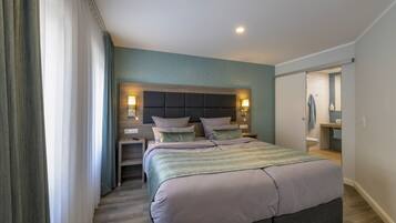 Quarto de hotel moderno com uma cama grande, uma mesa de cabeceira, uma luminária fixada na parede e um banheiro visível através de uma porta aberta.