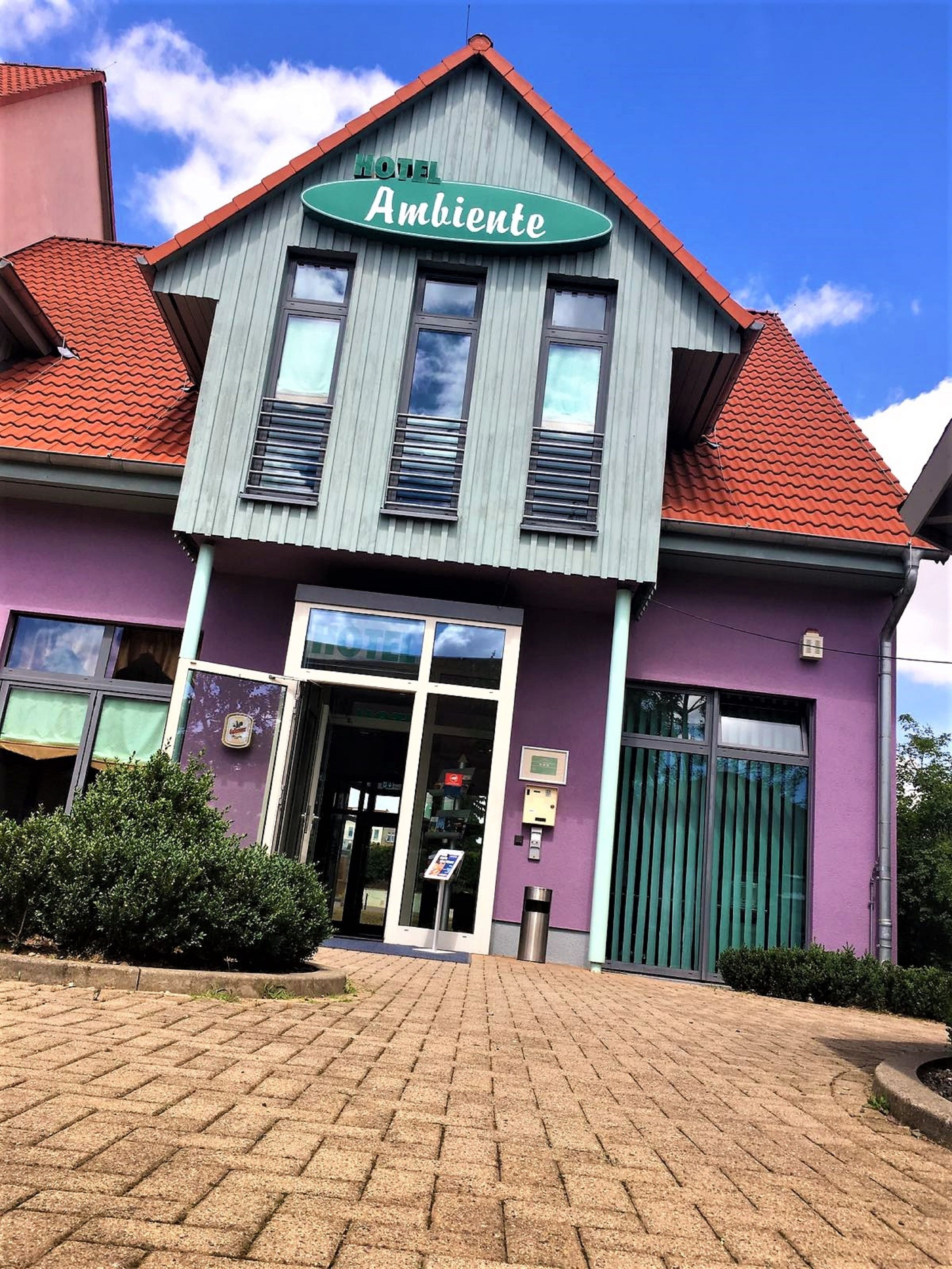 Um hotel chamado "Hotel Ambiente" com telhado de telhas vermelhas, fachada roxa e entrada com piso de tijolos.
